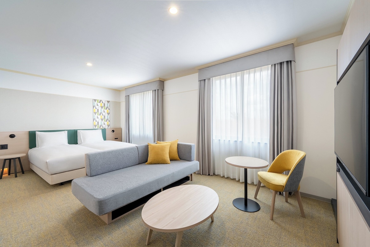 Quarto de hotel com cama, sofá, duas cadeiras, uma mesinha e televisão.