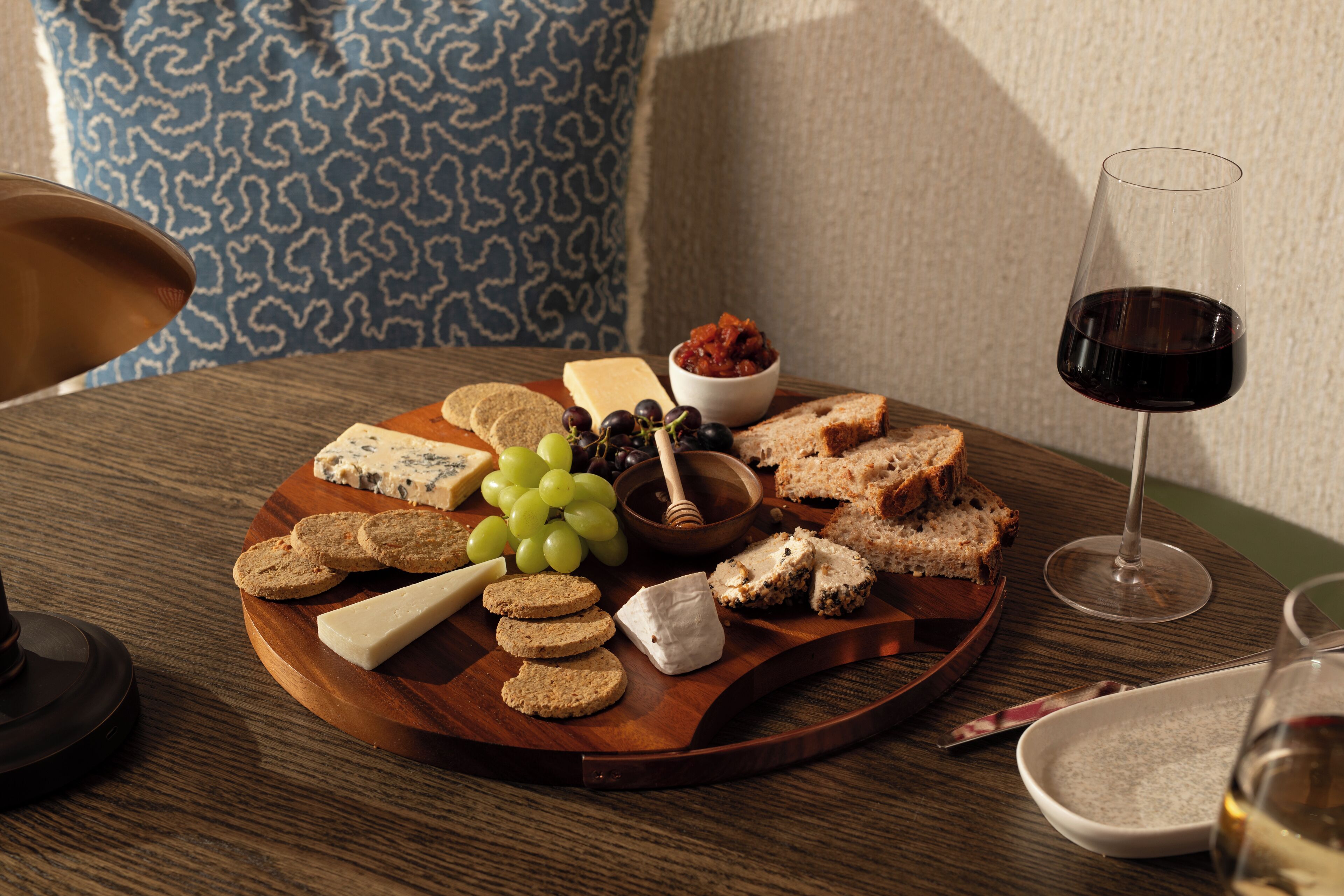 Tábua de madeira com queijo, uvas, bolachas e pão, acompanhada por um copo de vinho tinto.