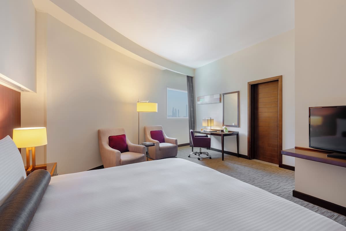 Quarto de hotel com uma cama grande, duas poltronas, uma escrivaninha, televisão e vista para a cidade.