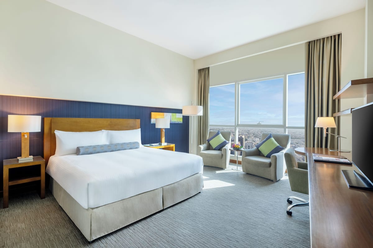 Quarto de hotel com uma cama grande, duas mesas de cabeceira com luminárias, uma escrivaninha, uma cadeira e vista para a cidade.