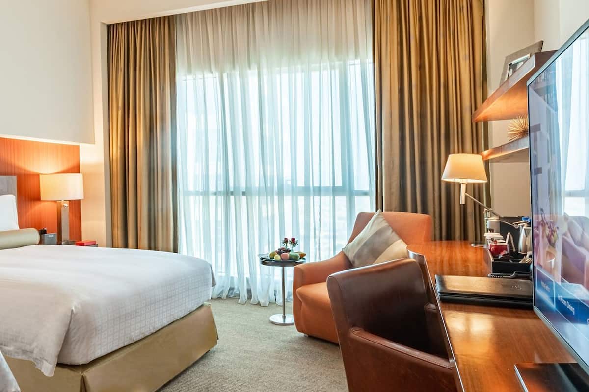 Quarto de hotel com cama, escrivaninha, cadeira, televisão e janela com cortinas.