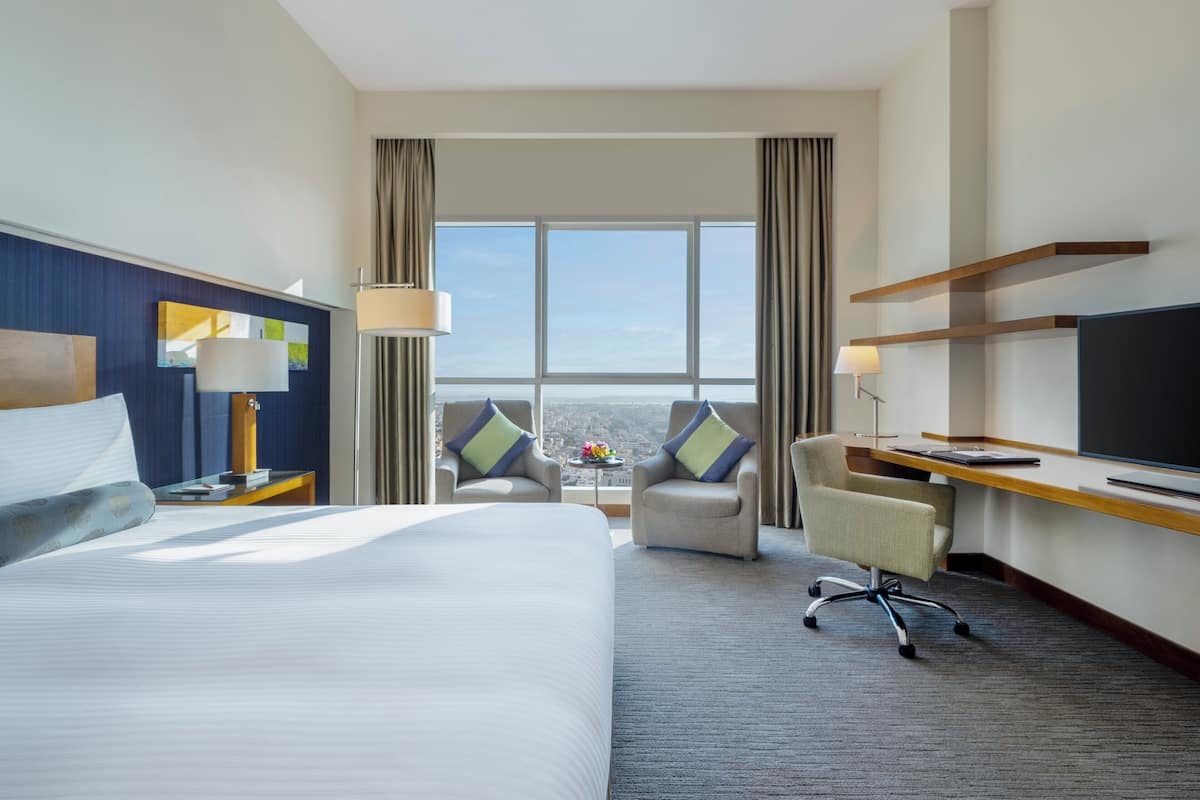 Quarto de hotel com uma cama grande, uma escrivaninha com computador, uma cadeira e uma televisão. Há uma janela com vista para a cidade.