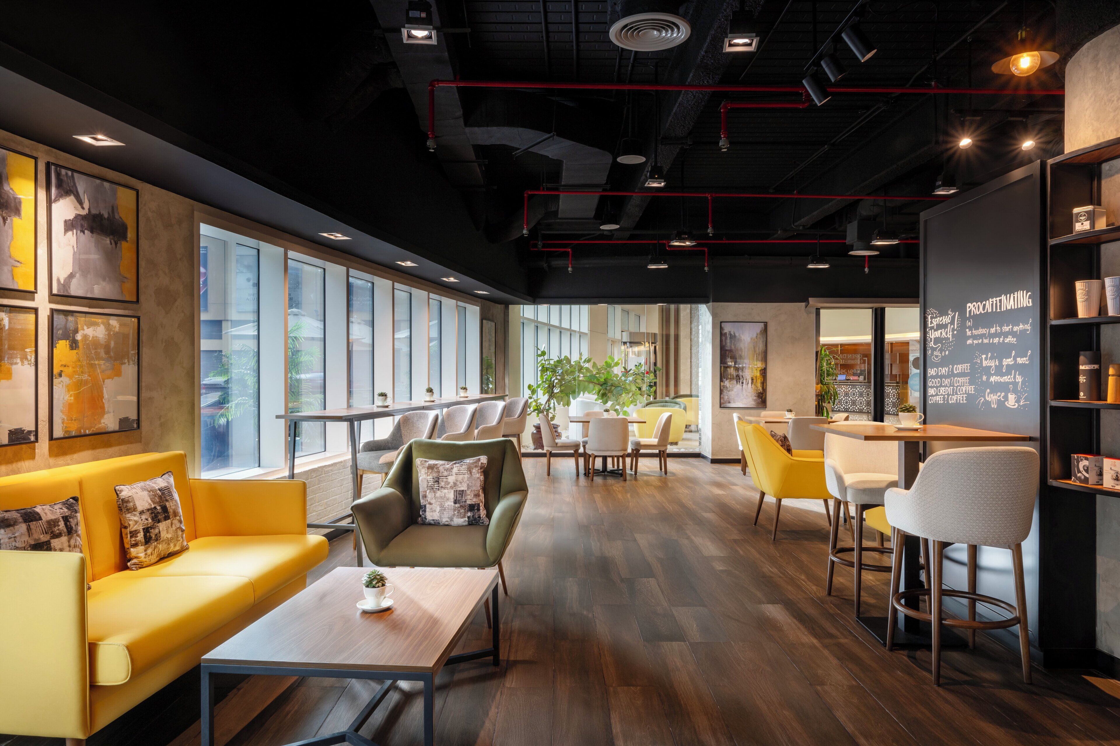 Um café moderno com piso de madeira, assentos em amarelo e verde, e amplas janelas que proporcionam luz natural.