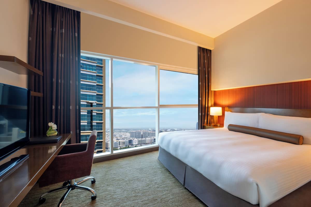 Quarto de hotel com uma cama grande, uma mesa com televisão, uma cadeira e vista para a cidade através de janelas amplas.