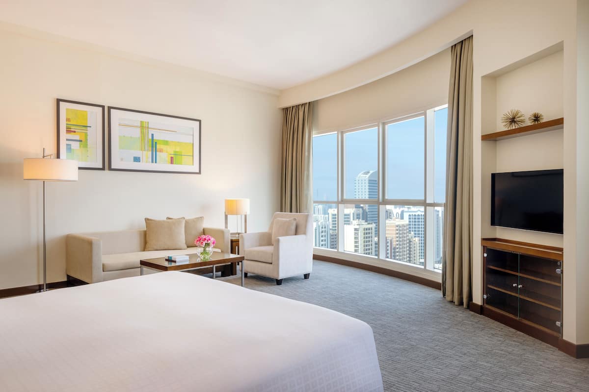 Quarto de hotel com uma cama grande, um sofá, uma cadeira, uma TV e uma janela ampla com vista para a cidade.