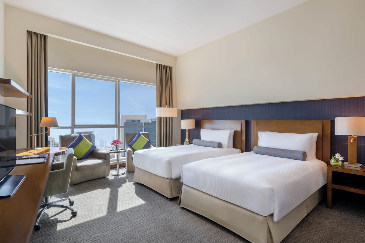 Quarto de hotel com duas camas, uma escrivaninha, uma cadeira, um abajur e vista para a cidade.
