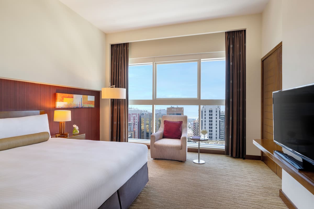 Quarto de hotel com uma cama grande, uma cadeira, uma televisão, uma janela com vista para a cidade e um abajur.