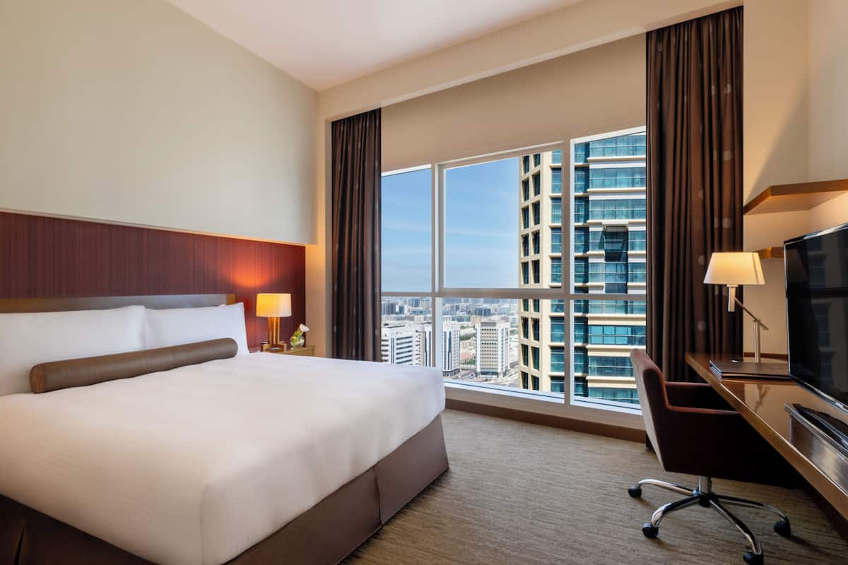 Quarto de hotel com uma cama grande, uma escrivaninha com televisão e vista para a cidade através de amplas janelas.