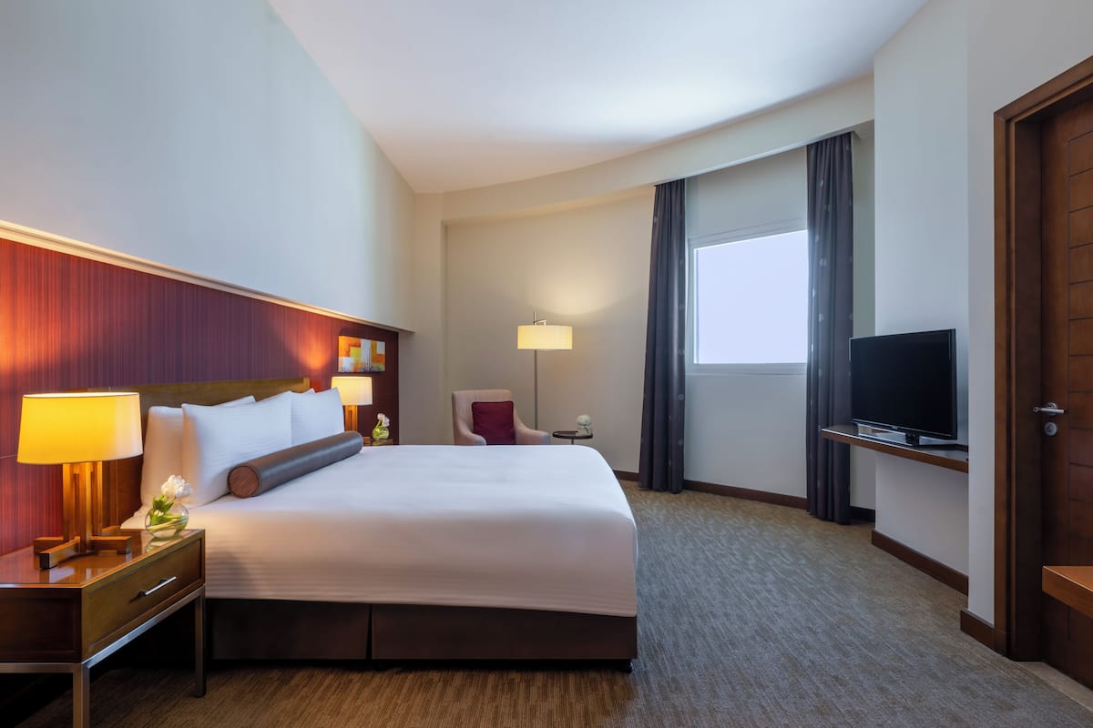 Quarto de hotel com uma cama grande, abajures, televisão, uma cadeira e uma janela com cortinas.