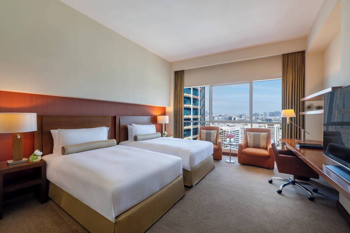 Quarto de hotel com duas camas, uma mesa com televisão, uma cadeira e vista da cidade através de grandes janelas.