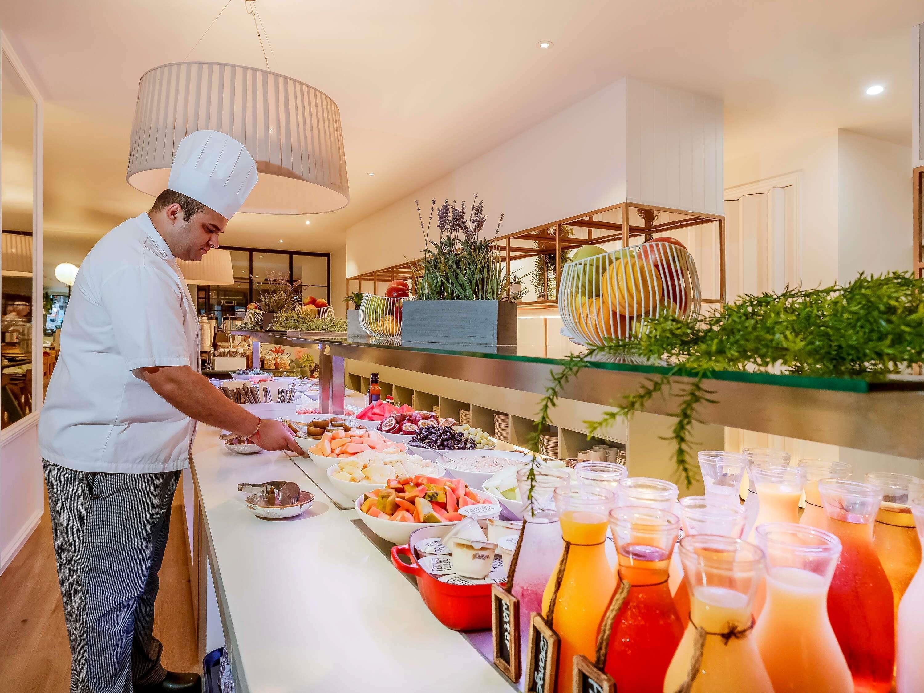 Buffet com diversos alimentos e bebidas, um chef de cozinha uniformizado e decoração de interiores moderna.