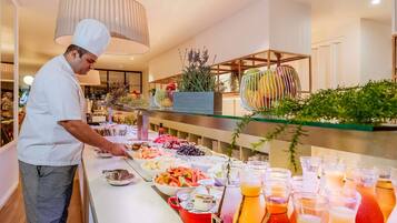 Buffet com diversos alimentos e bebidas, um chef de cozinha uniformizado e decoração de interiores moderna.