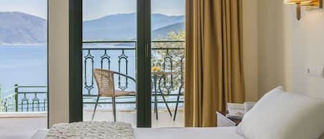 Quarto de hotel com cama, mesa de cabeceira, luminária, janela com vista para o mar e montanhas, e varanda com cadeira e mesa.