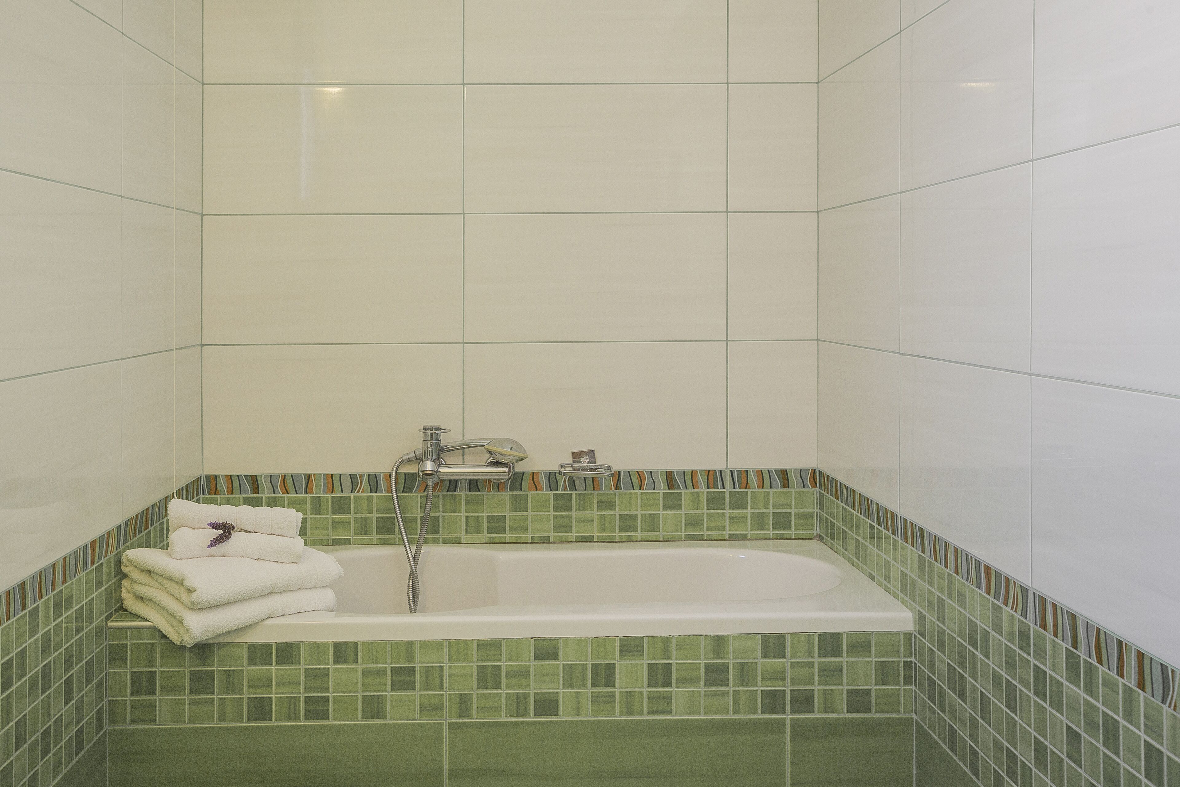 Um banheiro com banheira branca, azulejos de mosaico verde e uma pilha de toalhas brancas.