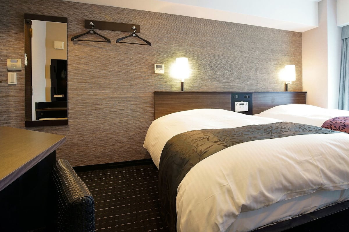 Quarto de hotel com duas camas, uma escrivaninha, um espelho e luminária fixada na parede.