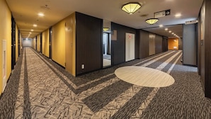 Corredor de hotel com carpete estampado, iluminação embutida e portas de acesso aos quartos.