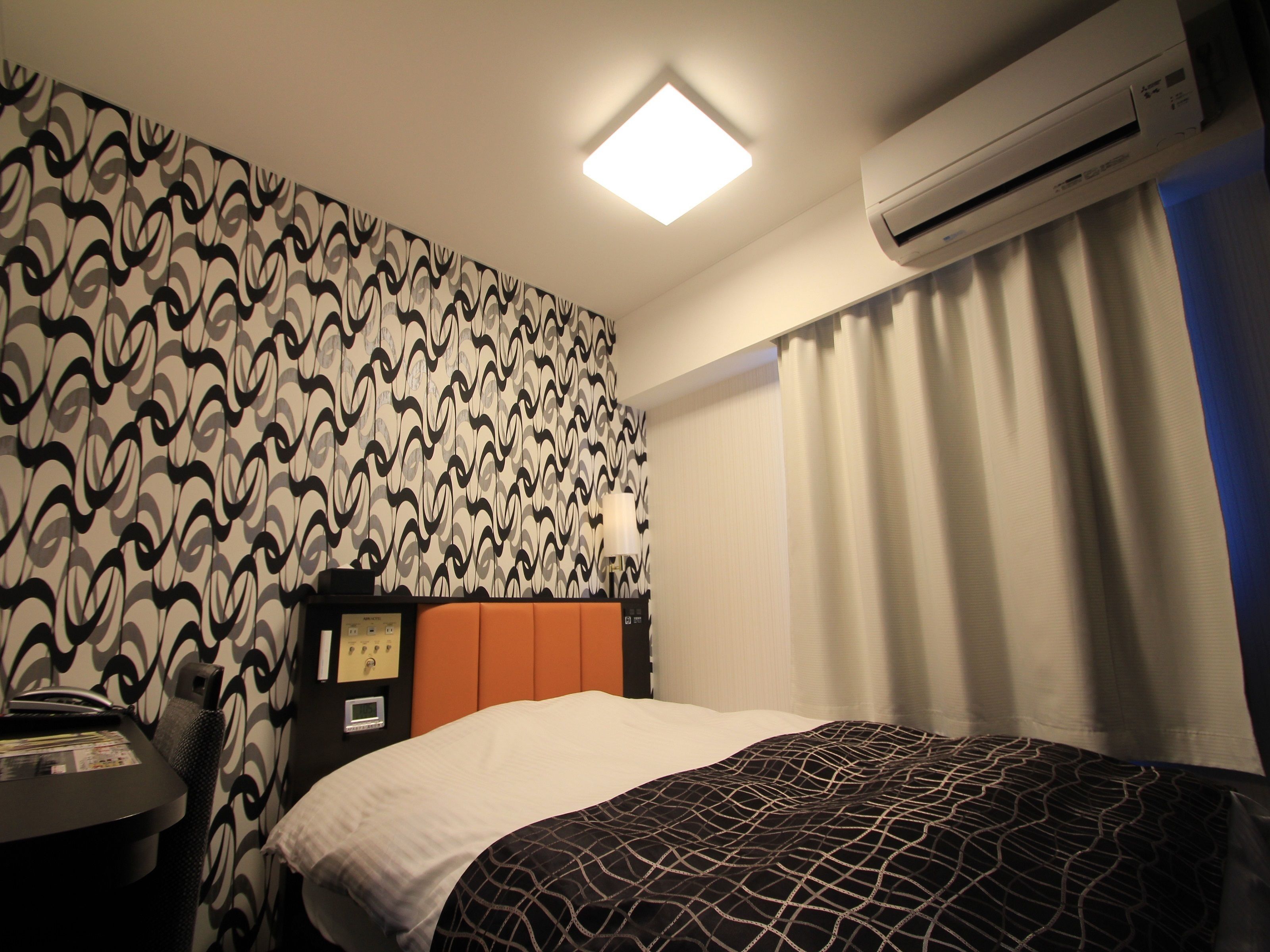 Quarto de hotel com papel de parede estampado, unidade de ar condicionado, cama com colcha estampada, mesa de cabeceira com abajur e janela com cortinas.