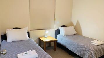 Quarto de hotel com duas camas de solteiro, cada uma com colcha listrada e travesseiros brancos. Há uma pequena mesa de cabeceira de madeira com uma luminária entre as camas. Uma toalha dobrada está sobre a cama à direita.