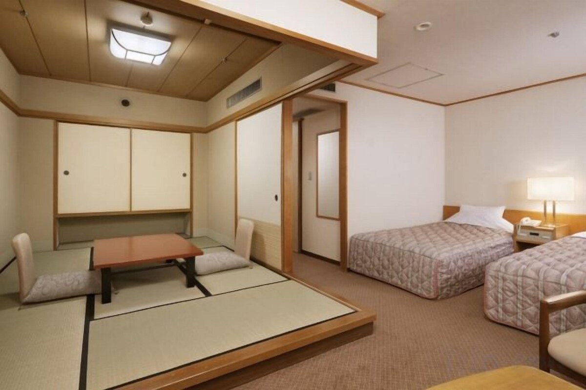 Quarto de hotel com piso de esteira de tatame, uma mesinha de madeira baixa, duas camas com roupa de cama estampada e uma mesa de cabeceira com abajur.