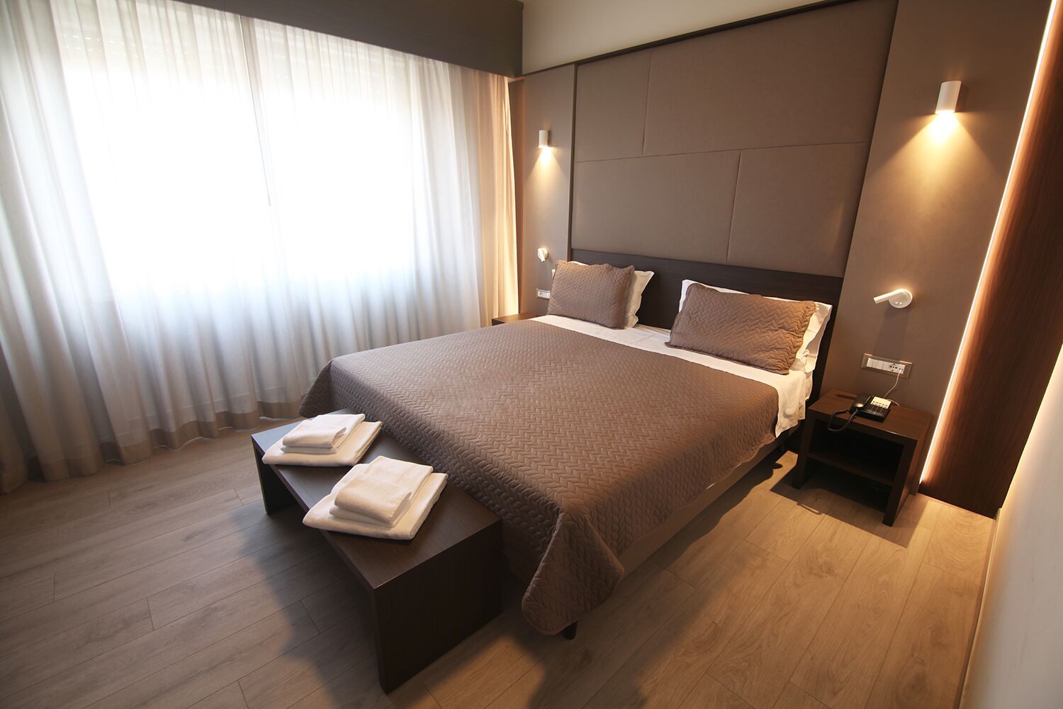 Quarto de hotel moderno com uma cama grande, uma mesinha de cabeceira com telefone e uma janela com cortinas leves.