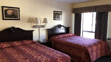 Quarto de hotel com duas camas, uma mesa de cabeceira com telefone e luminária, e uma janela com cortinas.