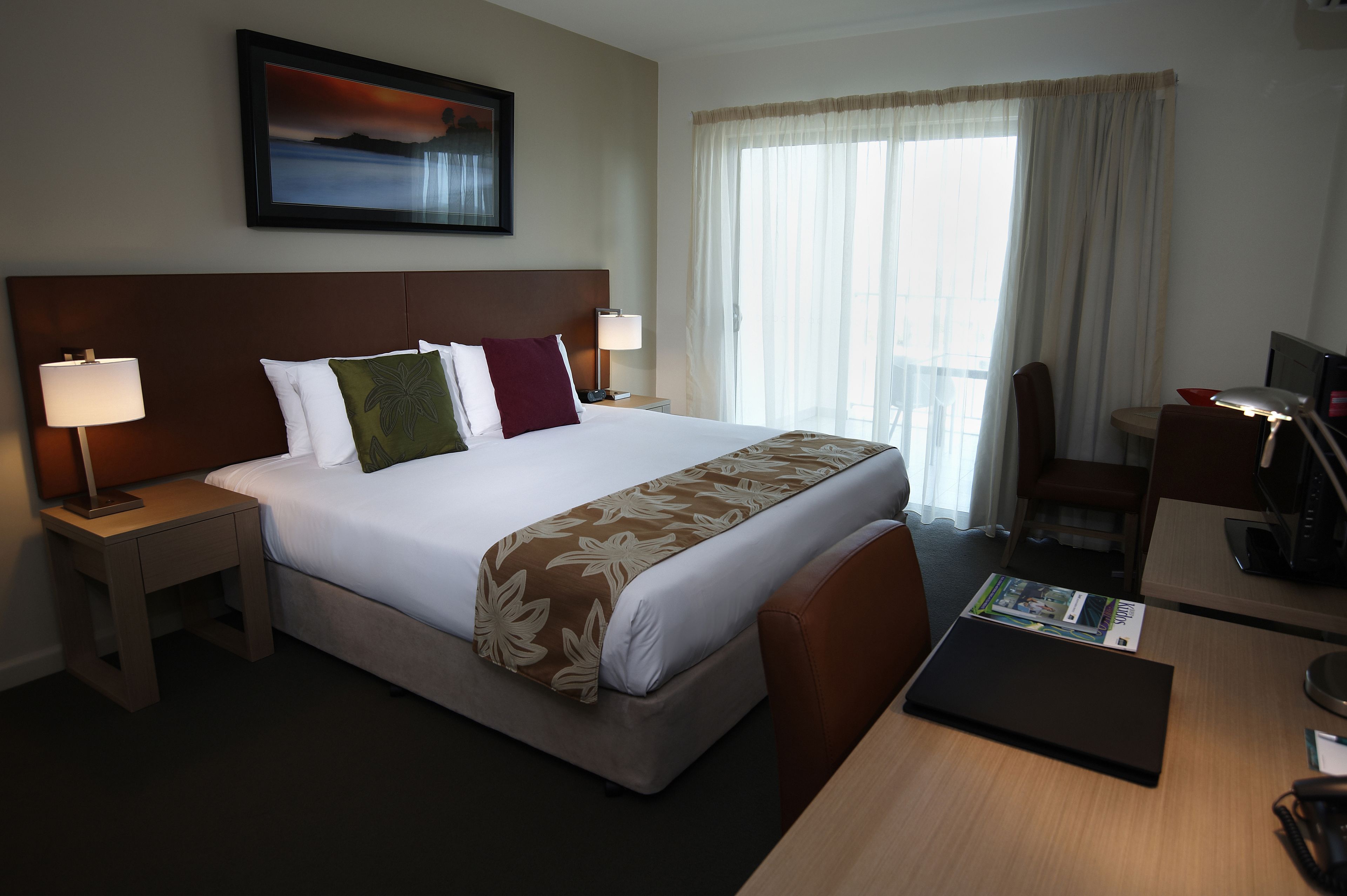 Quarto de hotel com uma cama grande, duas mesas de cabeceira com luminárias, uma escrivaninha com cadeira e uma janela com cortinas.