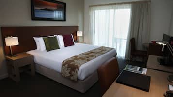Quarto de hotel com uma cama grande, duas mesas de cabeceira com luminárias, uma escrivaninha com cadeira e uma janela com cortinas.