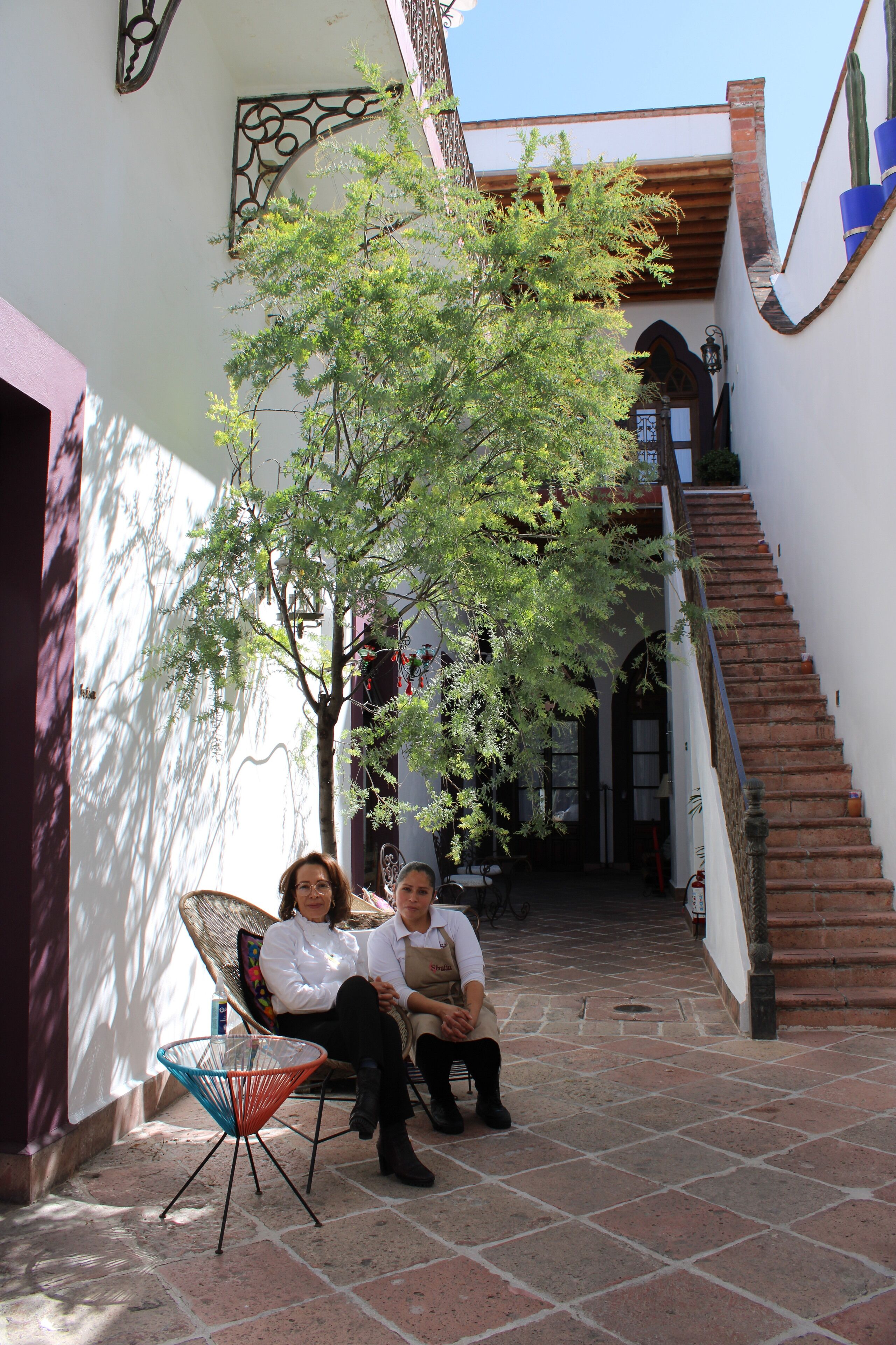 Um pátio estreito com uma escadaria, uma árvore e duas pessoas sentadas em cadeiras.
