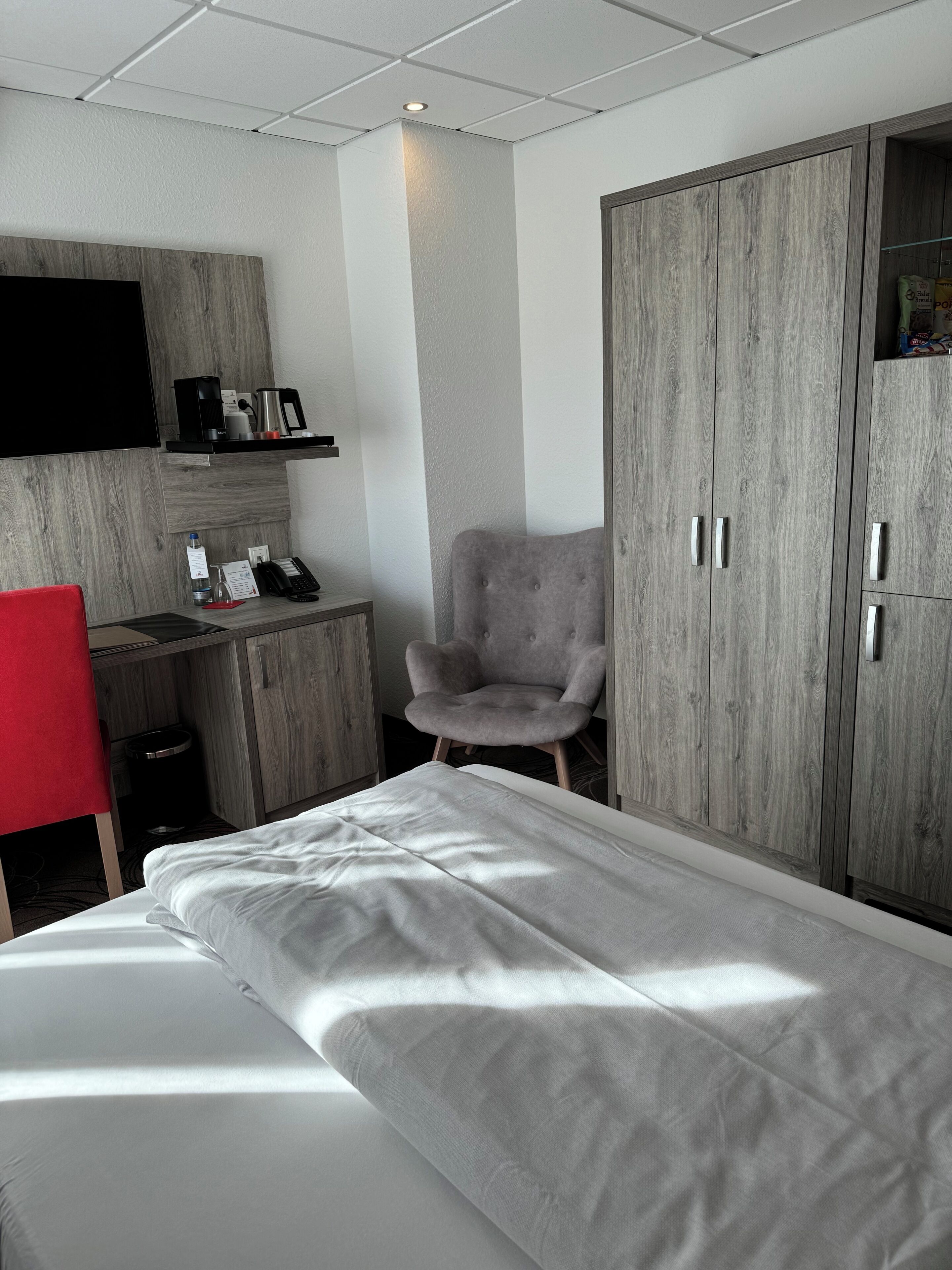 Quarto de hotel com cama, poltrona, guarda-roupa, secretária com máquina de café e televisão de ecrã plano na parede.