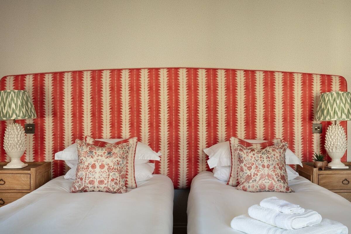 Quarto de hotel com duas camas, cada uma com almofadas estampadas com motivos florais, cabeceira listrada em vermelho e bege, e luminárias de cabeceira.