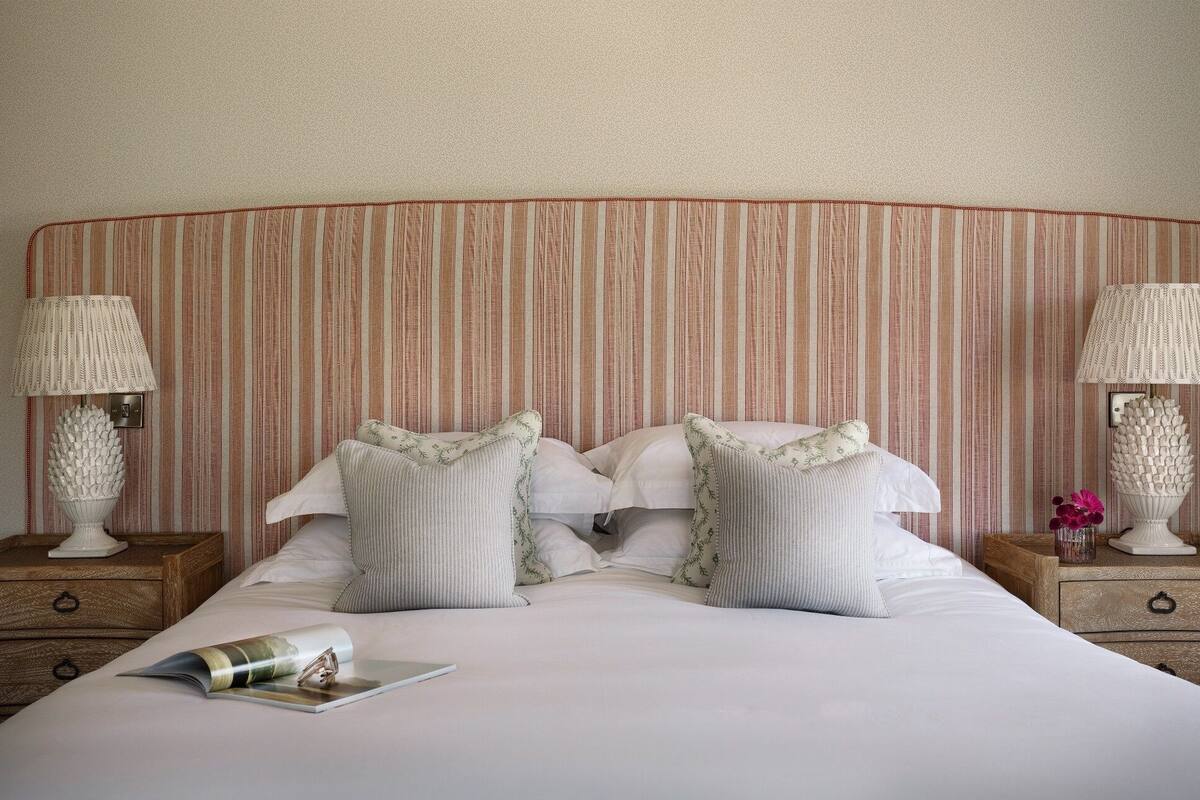 Cama bem arrumada com lençóis brancos e almofadas decorativas, com duas mesas de cabeceira com luminárias e uma pequena gaveta.