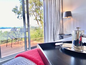 Quarto de hotel moderno com uma grande porta de vidro deslizante que dá para um deck externo, uma cama com detalhe em vermelho, uma cafeteira e um abajur.