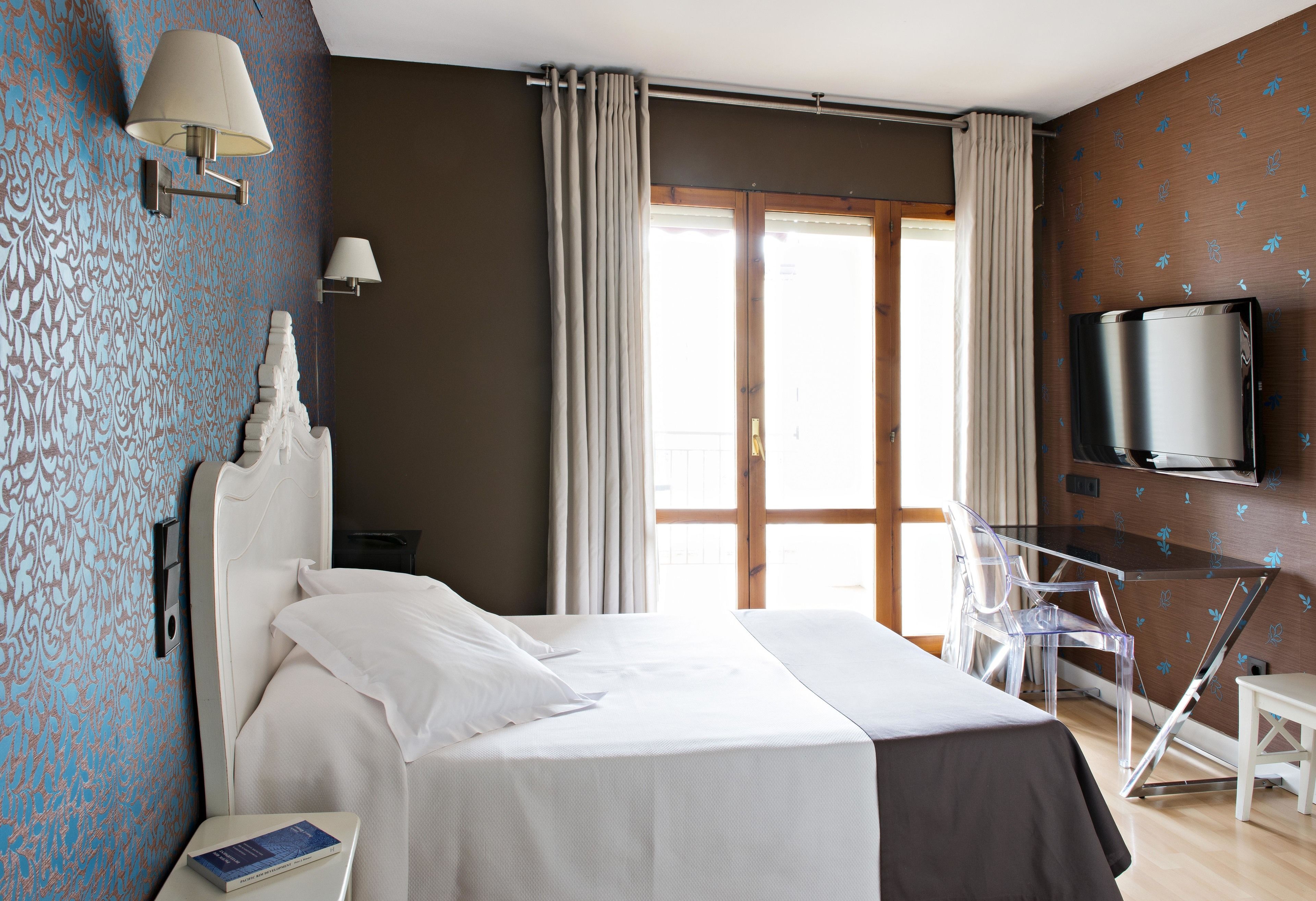 Quarto de hotel com cama, televisão, escrivaninha com cadeira e janela com cortinas.