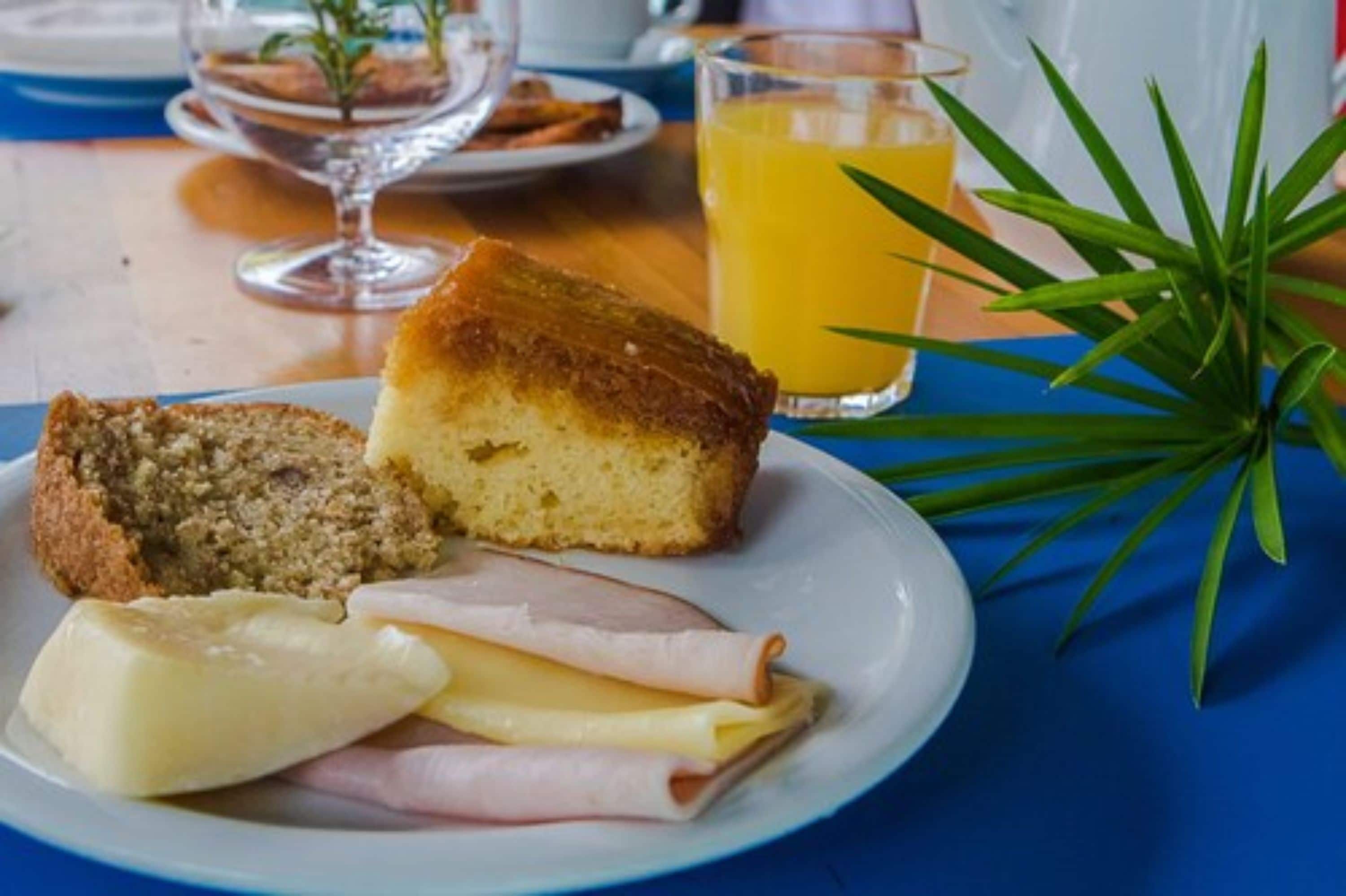 Prato de café da manhã com pão, queijo e presunto, um copo de suco de laranja e uma planta sobre uma mesa.