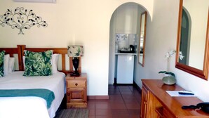 Quarto de hotel com cama, mesa de cabeceira, abajur, cômoda, espelho e uma cozinha compacta ao fundo.