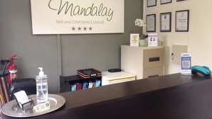Uma área de recepção com uma placa indicando "Mandalay B&B and Conference Centre", um extintor de incêndio, um dispensador de água, uma mesa com uma bandeja, um scanner e certificados emoldurados na parede.