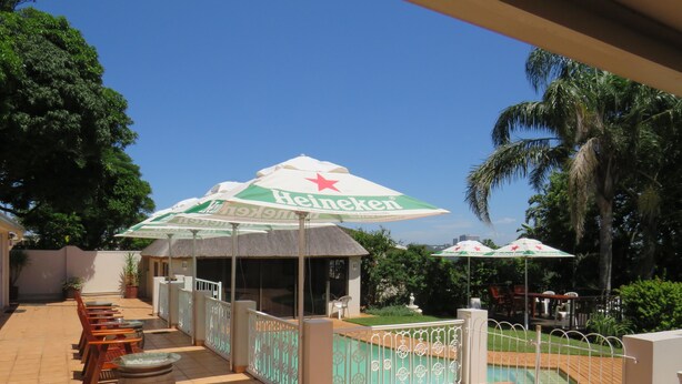 Uma área externa com mesas e guarda-sóis da marca Heineken, piscina e uma estrutura com telhado de palha.