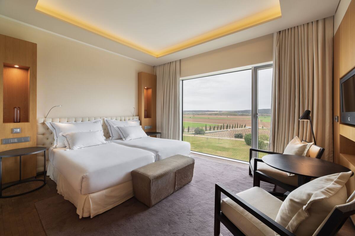 Quarto de hotel com uma cama grande, uma cama menor, um sofá, uma escrivaninha e uma janela ampla com vista para o campo.