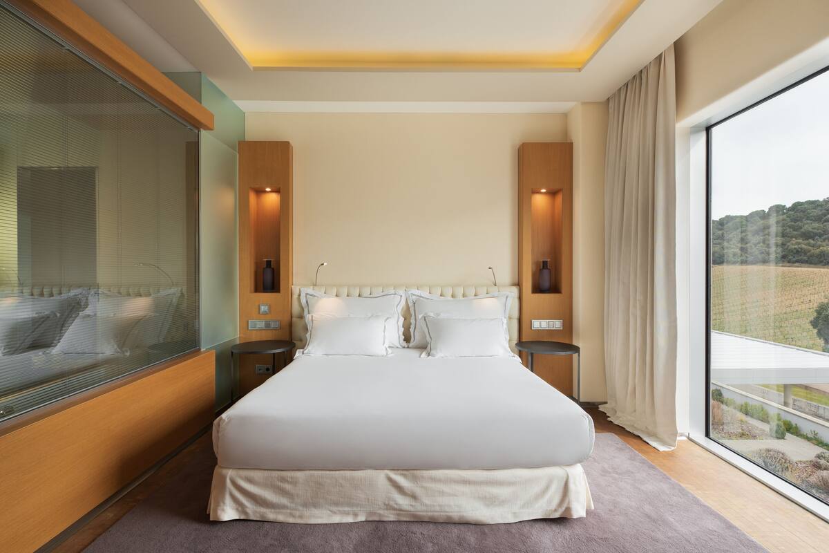 Uma cama grande com lençóis brancos, uma divisória de vidro e vista para o exterior.