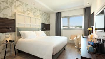 Quarto de hotel com uma cama grande, uma escrivaninha com cafeteira, uma cadeira e vista para a cidade.