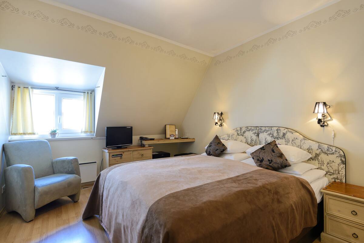 Quarto com cama, poltrona, escrivaninha com computador e janela com cortinas.