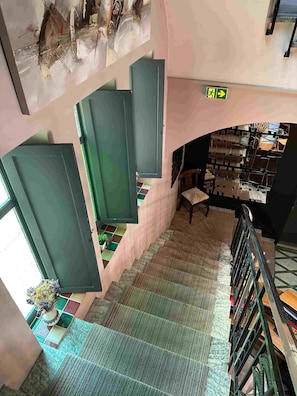 Uma escadaria com carpete estampado conduz a um andar inferior, uma porta verde e uma parede com painéis quadrados dispostos em grade.