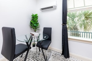 Quarto com mesa de vidro, duas cadeiras pretas, uma planta em vaso, ar condicionado branco e janela com vista para palmeiras.