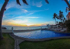 Piscina com vista para o pôr do sol sobre o oceano, cercada por palmeiras e cadeiras de descanso.