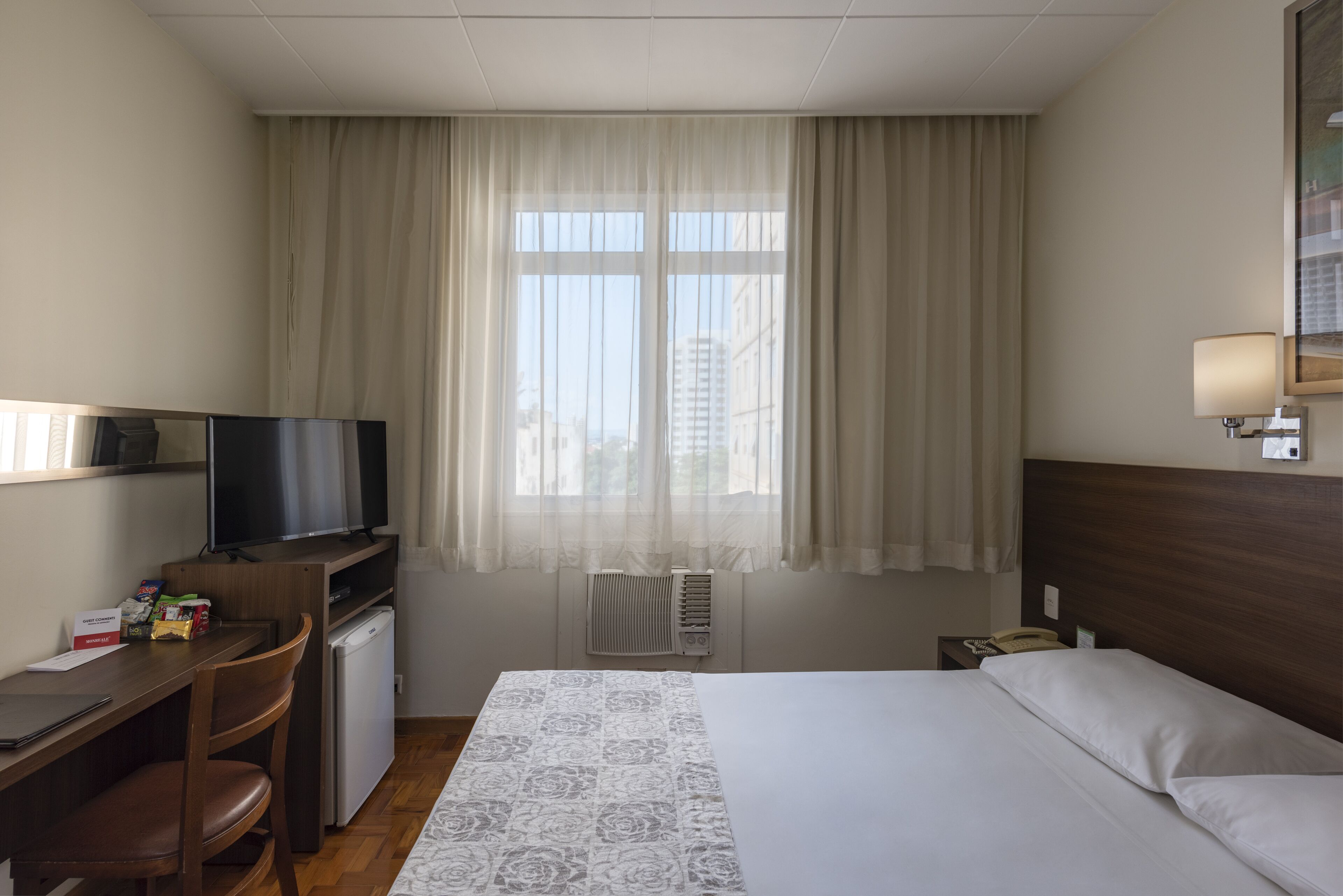 Quarto de hotel com uma cama, uma escrivaninha com computador, televisão e ar condicionado.