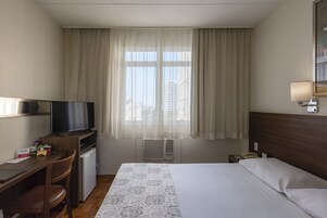 Quarto de hotel com uma cama, uma escrivaninha com computador, televisão e ar condicionado.