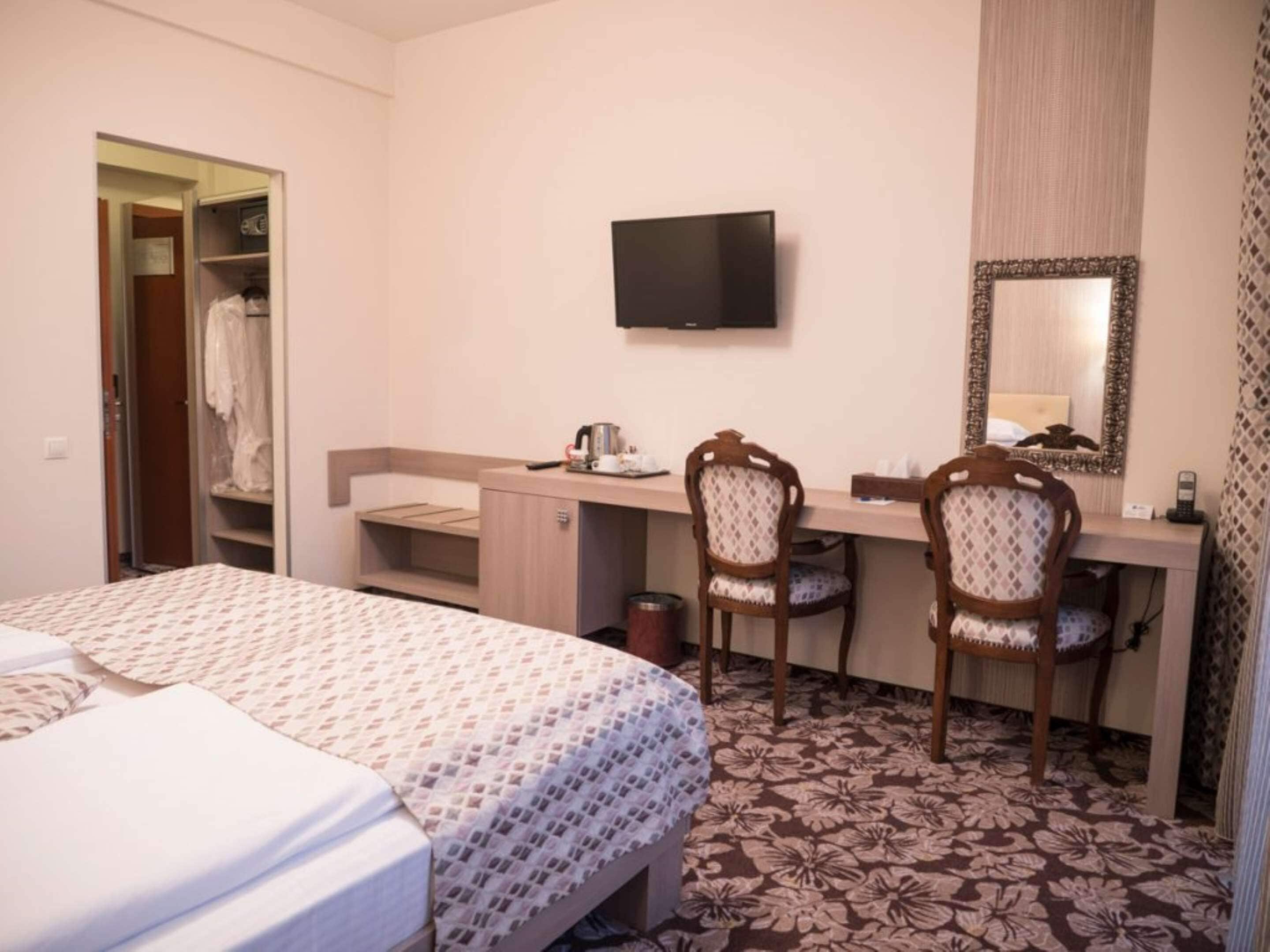 Quarto de hotel com cama, escrivaninha, duas cadeiras, televisão e espelho.