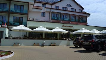 Um edifício com restaurante no andar térreo, mesas ao ar livre com guarda-sóis e carros estacionados.