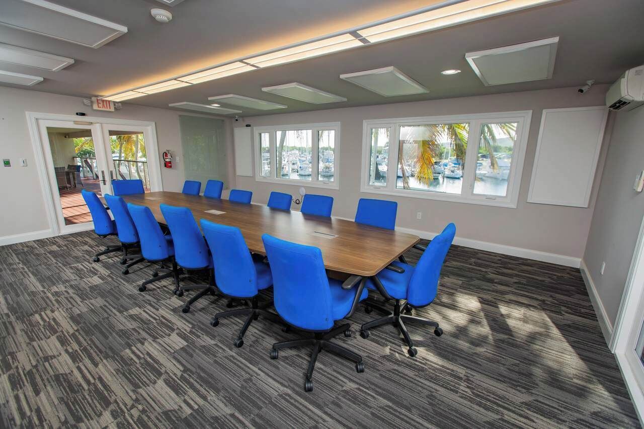 Uma sala de conferências com uma mesa comprida, cadeiras azuis e grandes janelas com vista para carros estacionados e palmeiras.