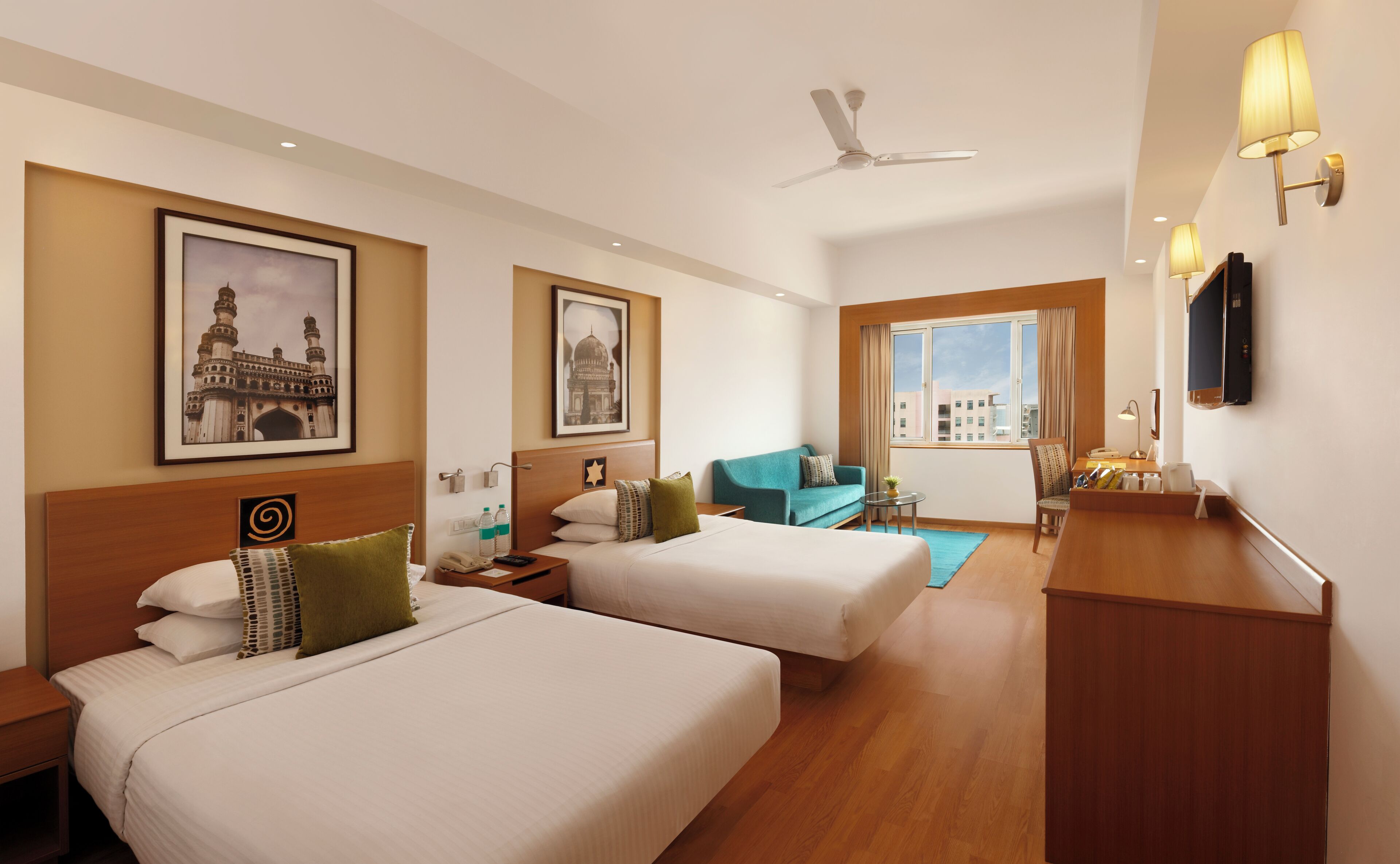 Quarto de hotel com duas camas, uma escrivaninha, uma cadeira, televisão e janela com vista para a cidade.